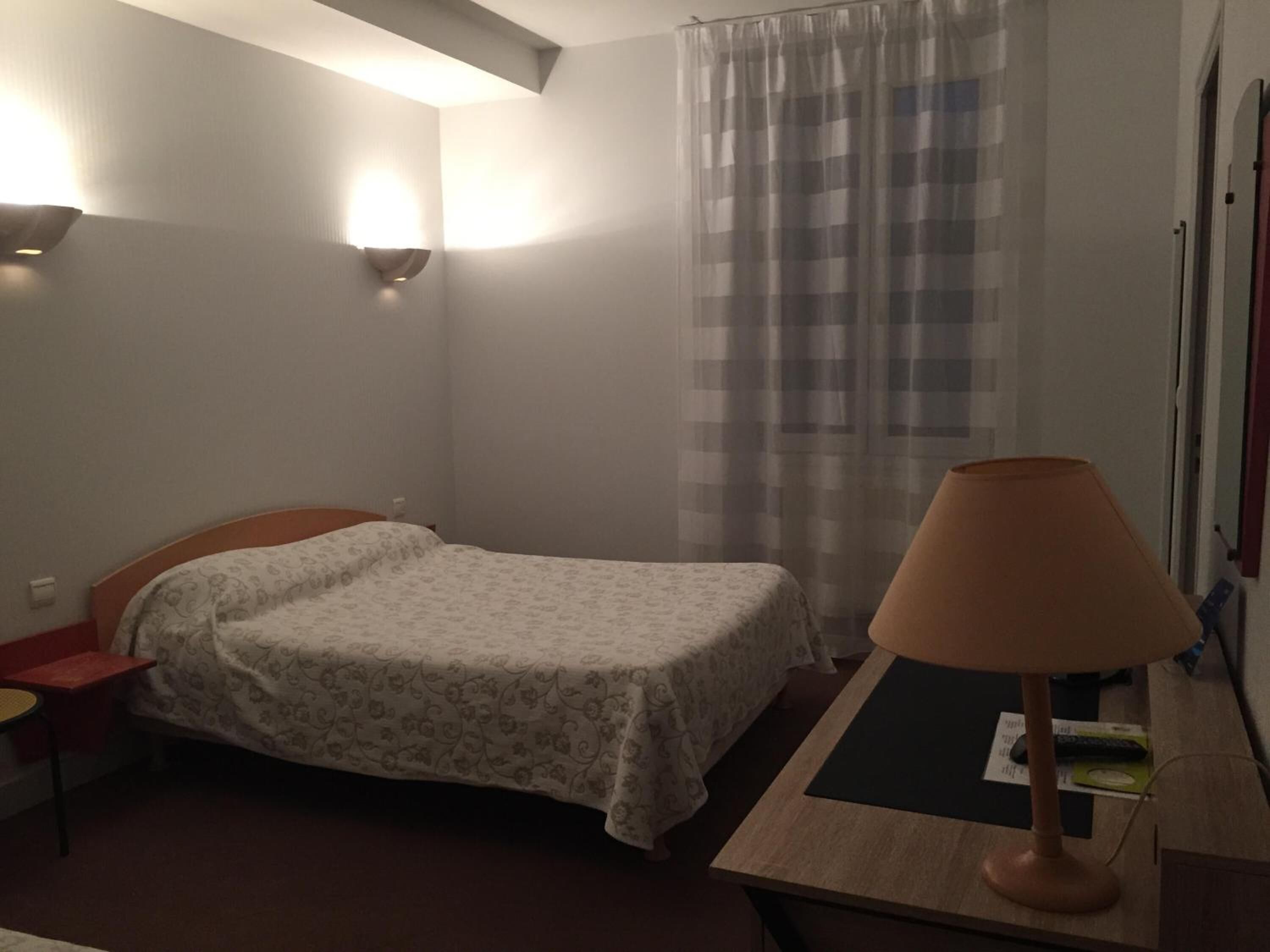 Quarto de hotel com uma cama, mesa de cabeceira, abajur e janela com cortinas.
