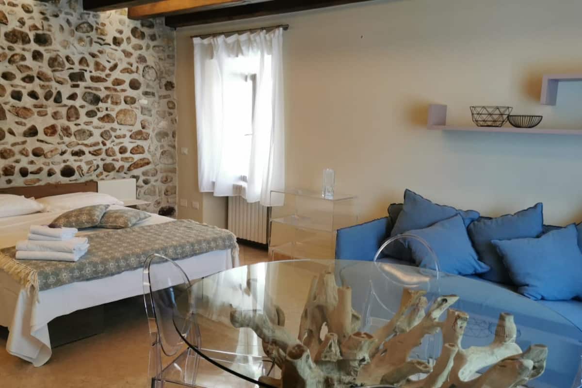 Quarto com parede de pedra, cama com colcha estampada, mesa de centro de vidro com escultura em madeira natural e sofá azul com almofadas a combinar.