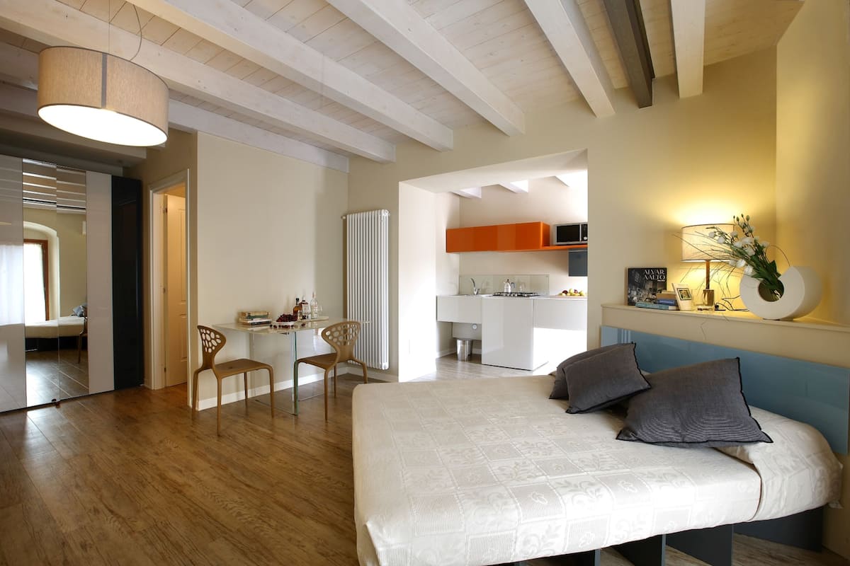 Quarto moderno com uma cama grande, uma escrivaninha e uma cadeira. É possível ver uma cozinha ao fundo, com armários laranja.