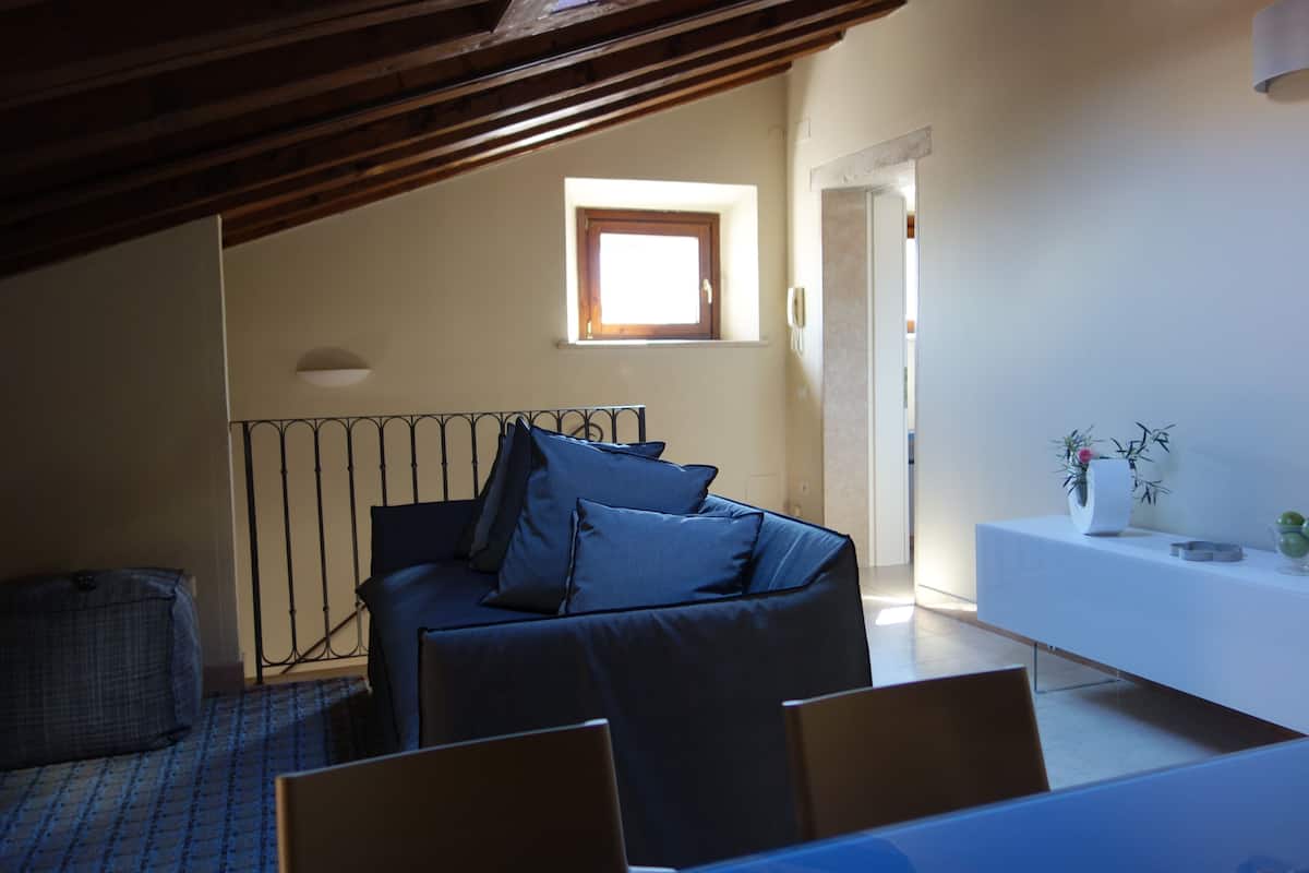 Quarto estilo loft aconchegante, com sofá azul escuro, uma pequena janela e uma mesinha de cabeceira branca com um vaso.