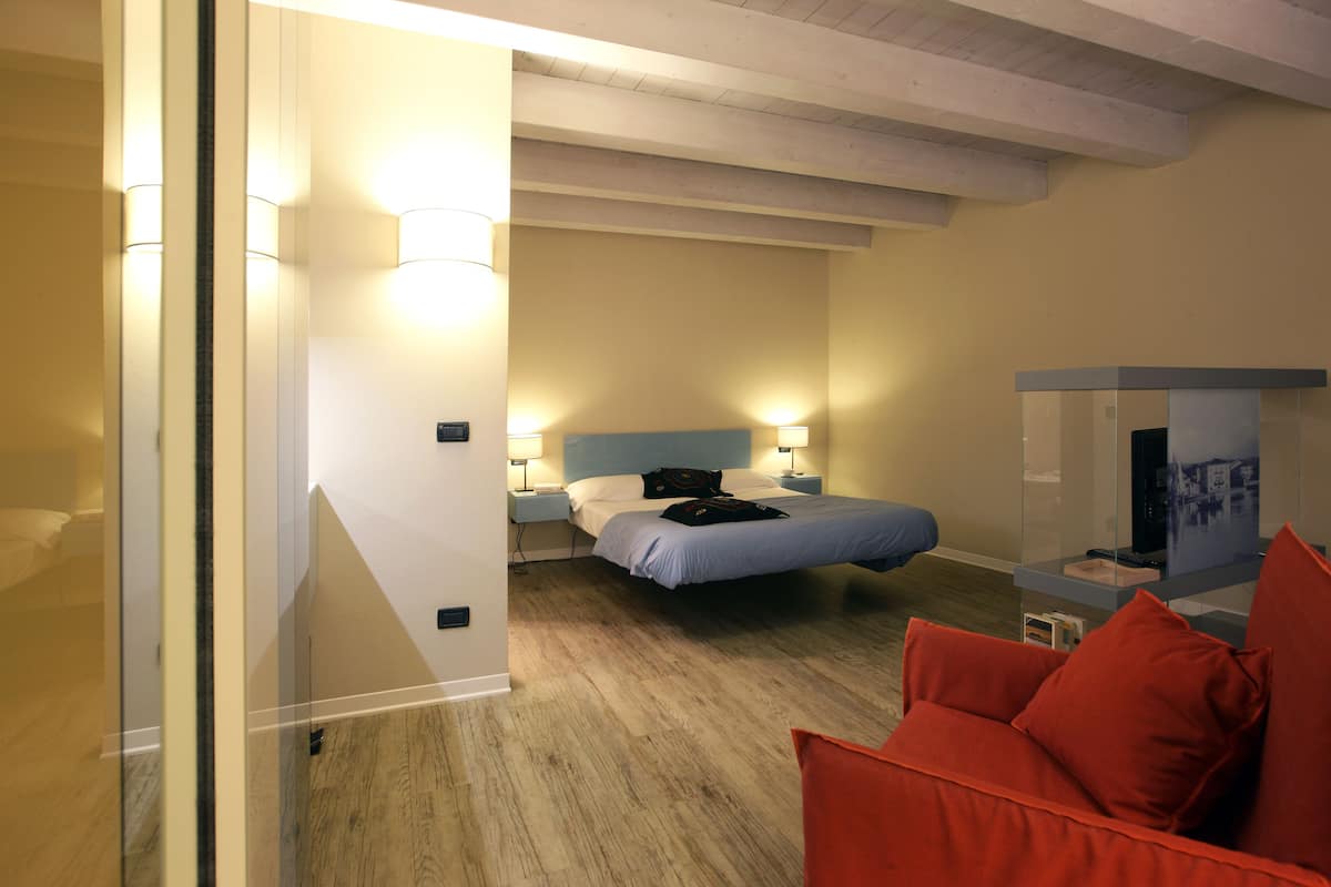 Quarto de hotel moderno com cama, mesas de cabeceira, uma cadeira vermelha e um painel de vidro.