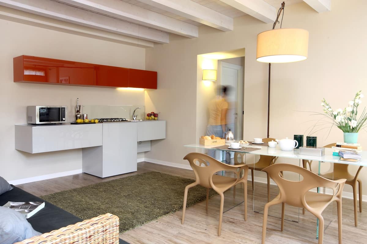 Cozinha moderna com armários brancos e um detalhe em vermelho, uma área de jantar com mesa de vidro e cadeiras bege, e uma luminária pendente grande.
