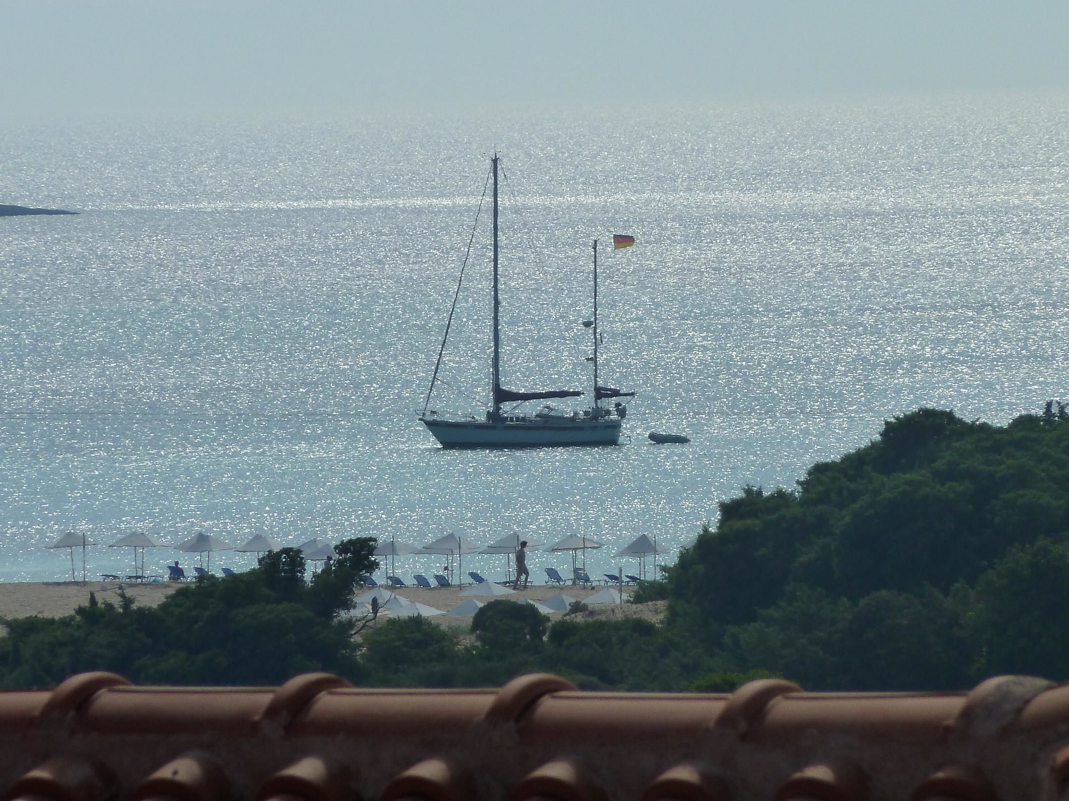 Um veleiro em um mar calmo, com uma praia e guarda-sóis ao fundo.