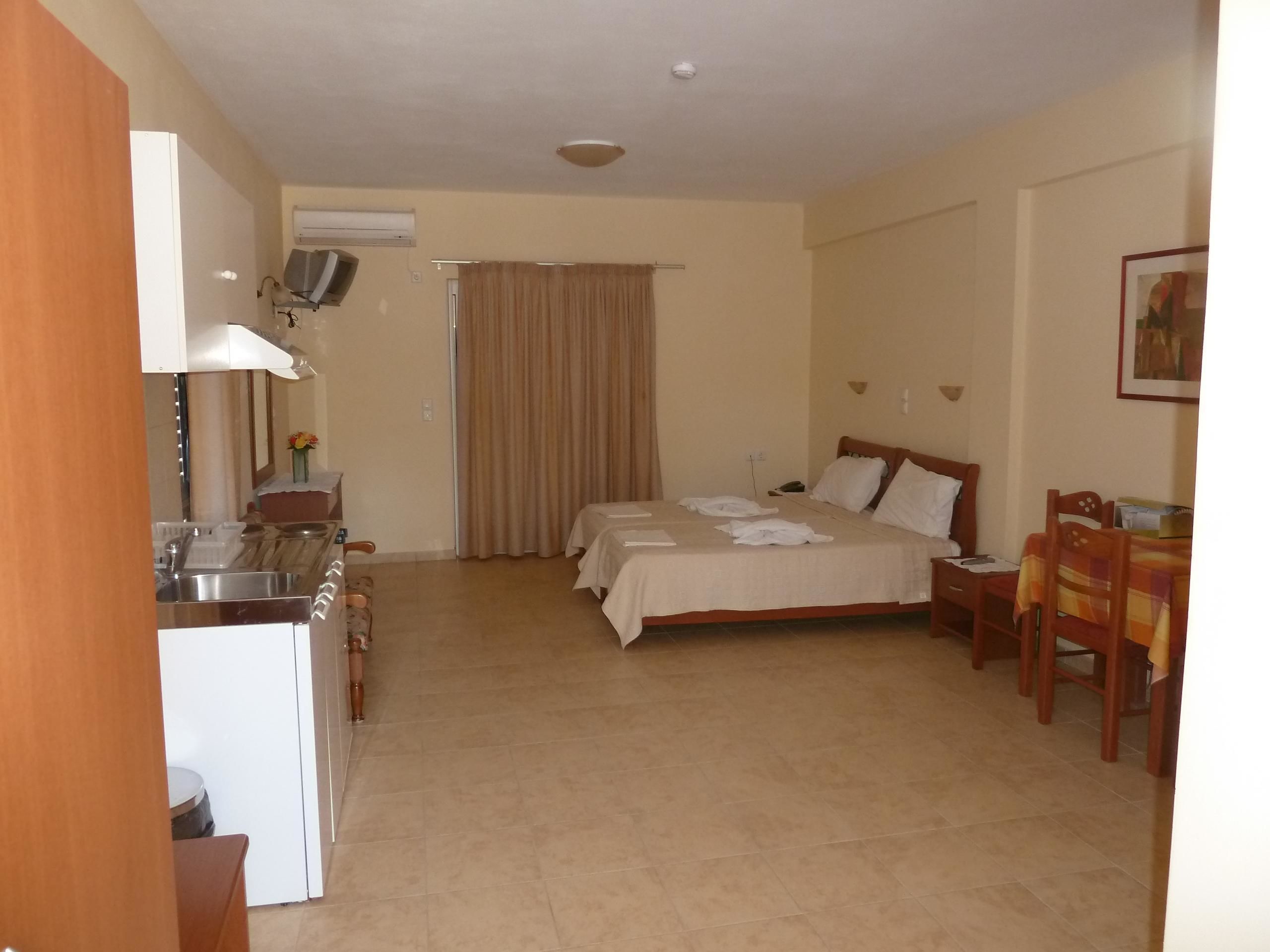 Quarto de hotel com cama, escrivaninha, cadeira, mesa pequena, espelho, janela com cortinas e cozinha compacta.