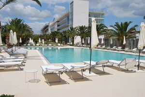 Uma área de piscina com espreguiçadeiras e guarda-sóis, cercada por palmeiras e edifícios modernos.