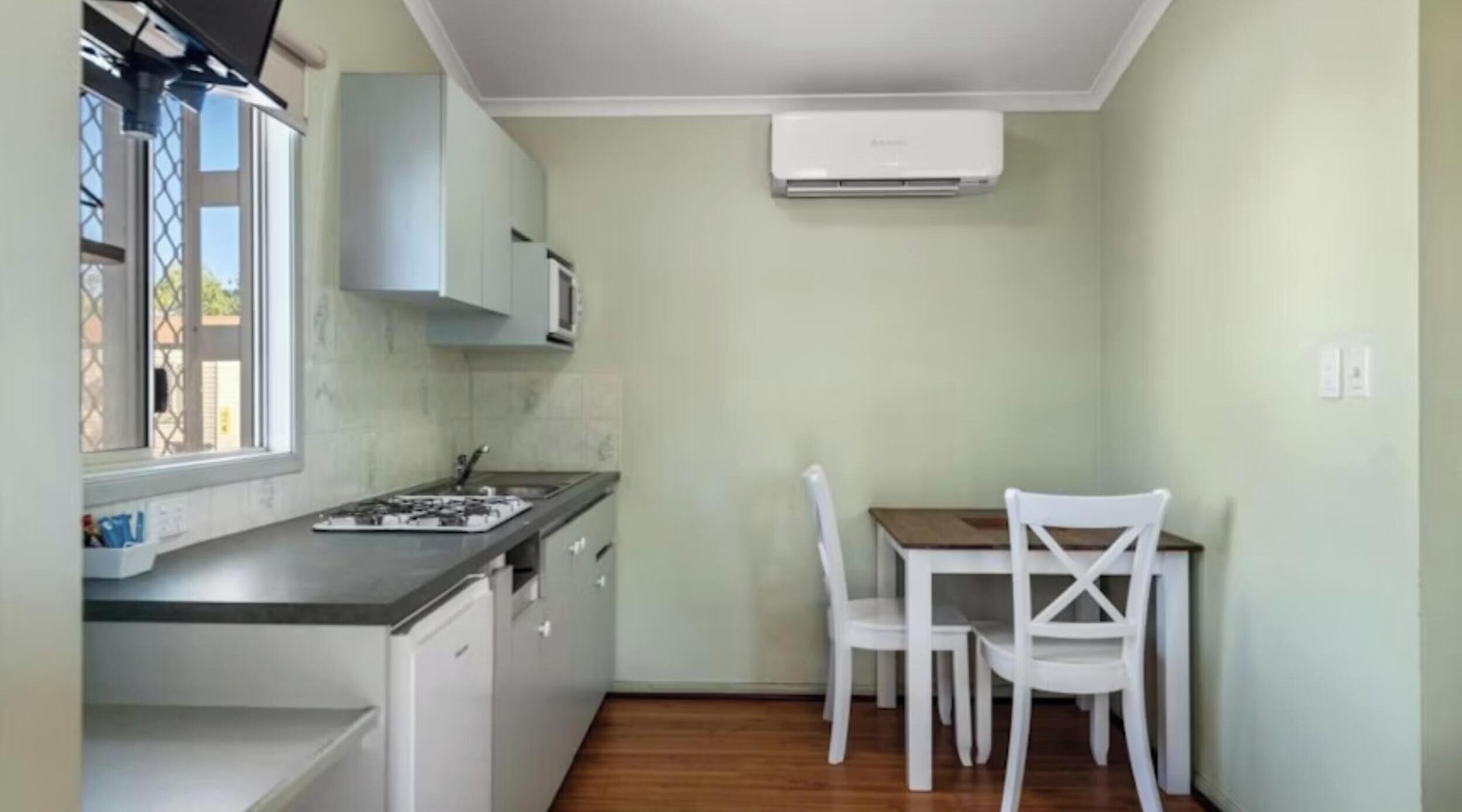Cozinha compacta com fogão a gás, armários brancos, mesa de jantar com duas cadeiras e um aparelho de ar condicionado fixado na parede.
