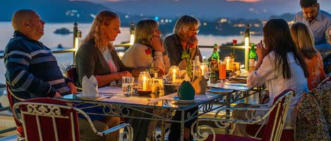 Um grupo de pessoas jantando em uma mesa ao ar livre, com vista panorâmica para montanhas e água ao fundo.
