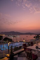 Terraço com piscina, cadeiras vermelhas e vista para a água ao entardecer.