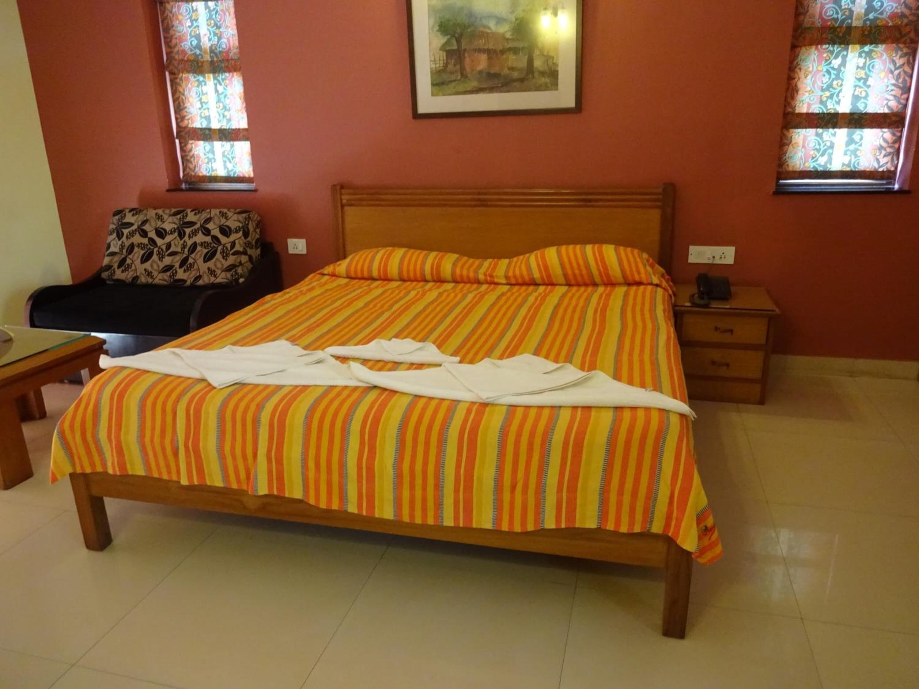 Quarto com cama de madeira, roupa de cama listrada, uma cadeira com almofada florida, mesa de cabeceira e um quadro na parede.