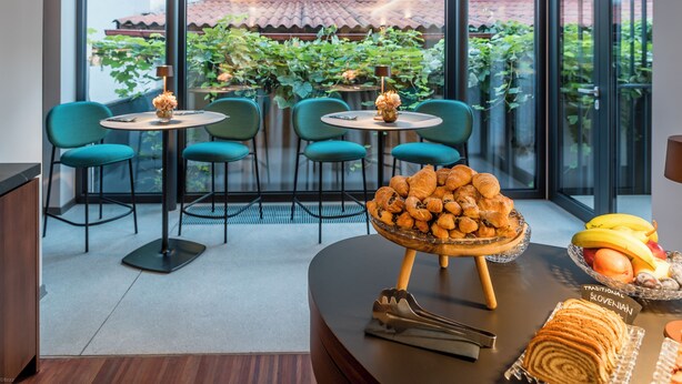Uma sala de jantar moderna com mesas redondas e cadeiras cor turquesa, um buffet com pastelaria e frutas, e vista para um edifício com telhado vermelho através de grandes janelas.