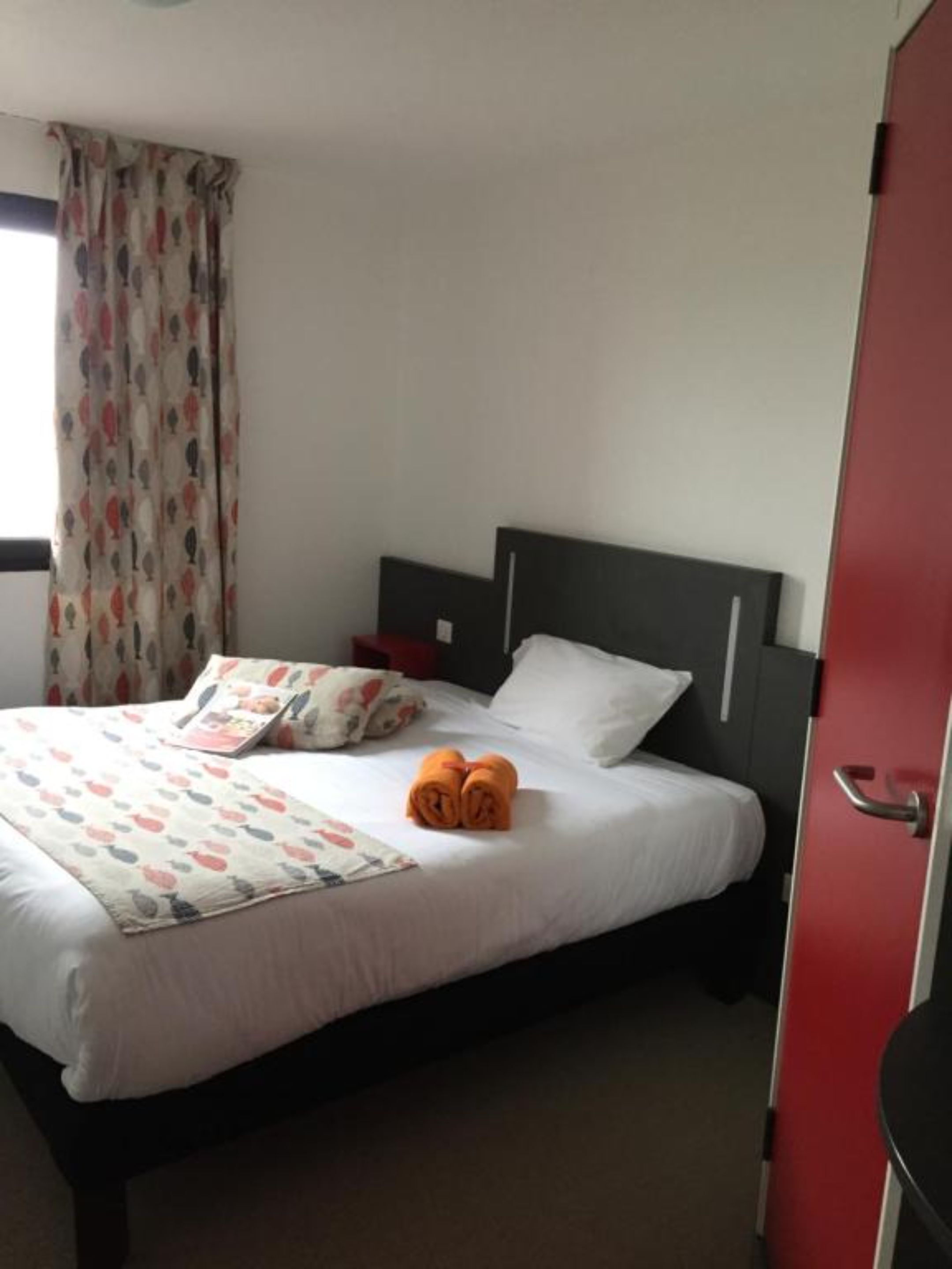Uma cama bem arrumada com colcha florida, um par de toalhas enroladas e uma janela com cortinas estampadas.