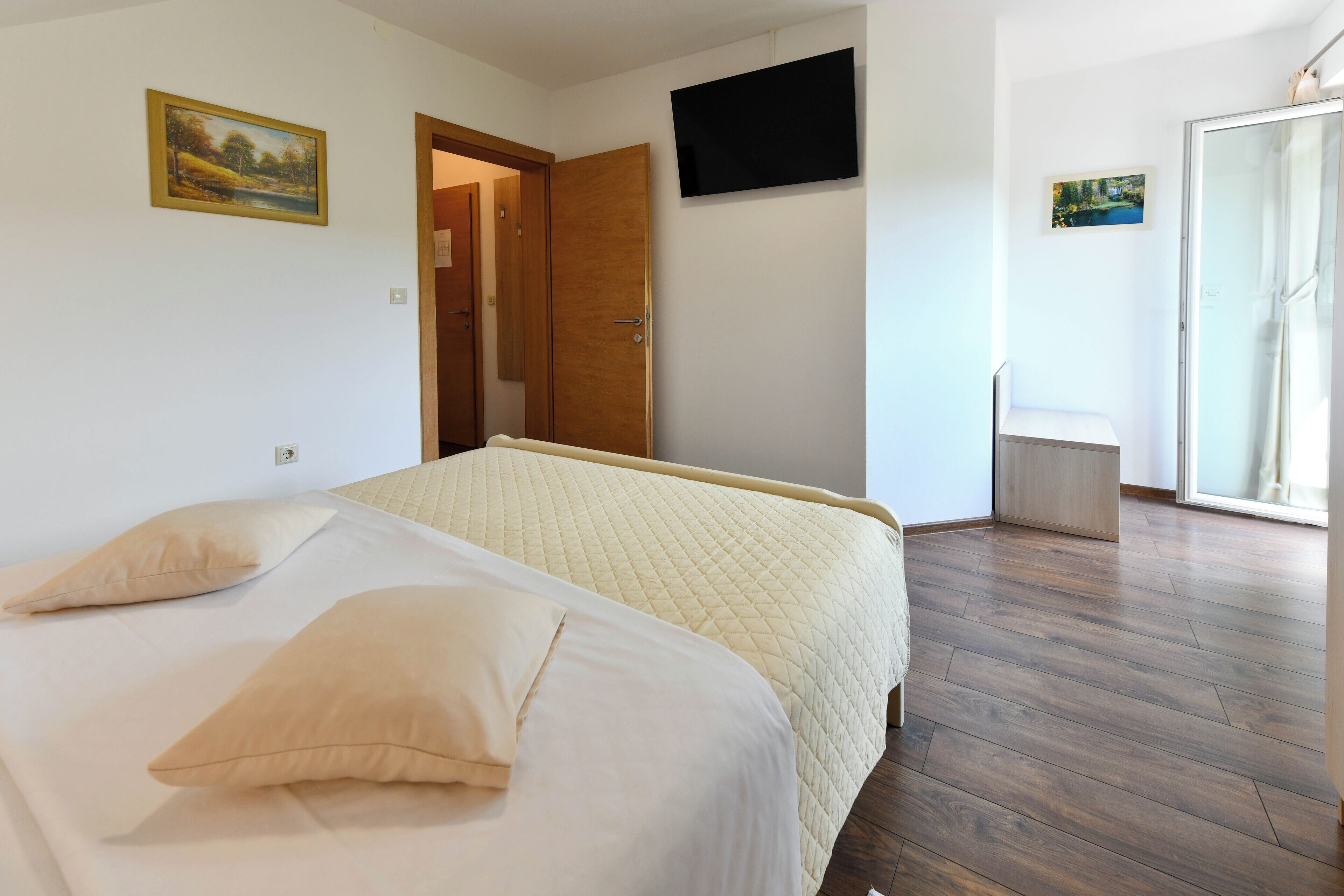 Um quarto com uma cama, uma porta de madeira, uma televisão e uma janela pequena.