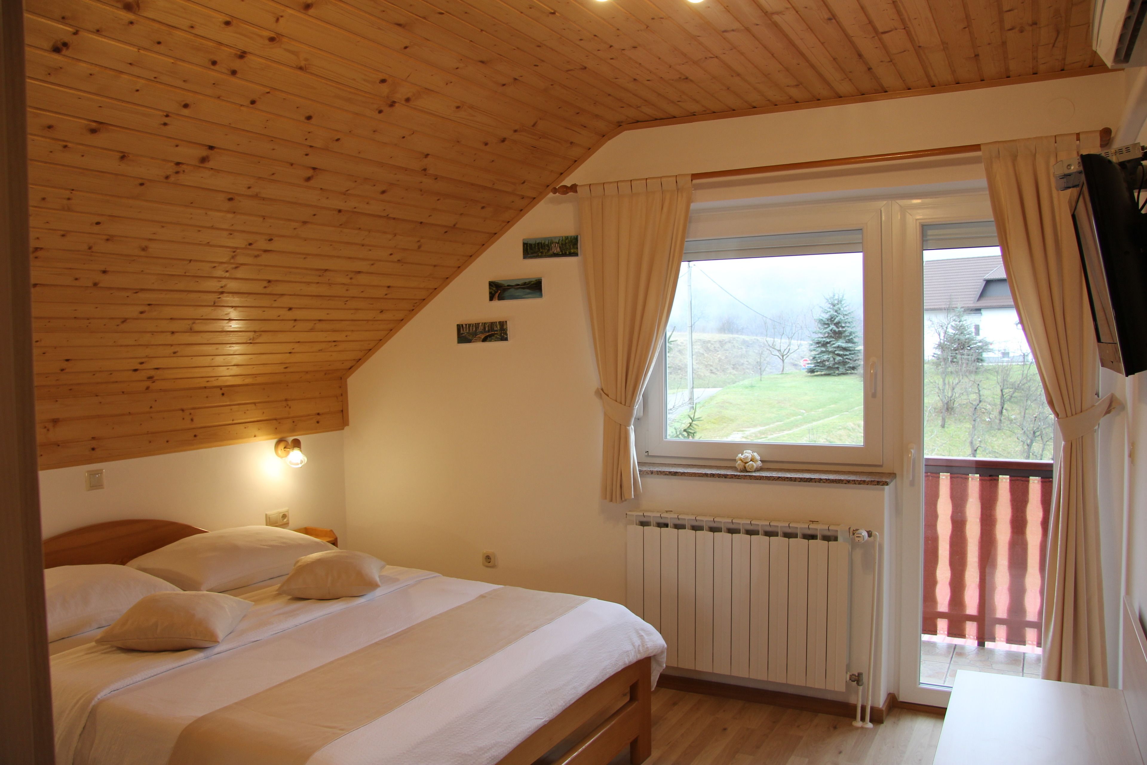 Quarto com teto inclinado, uma janela grande com cortinas, radiador e cama de madeira.