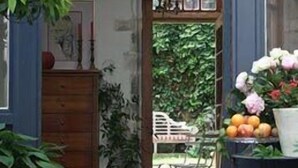 Porta de entrada para um jardim com um banco de madeira, plantas em vasos e uma mesa com frutas.