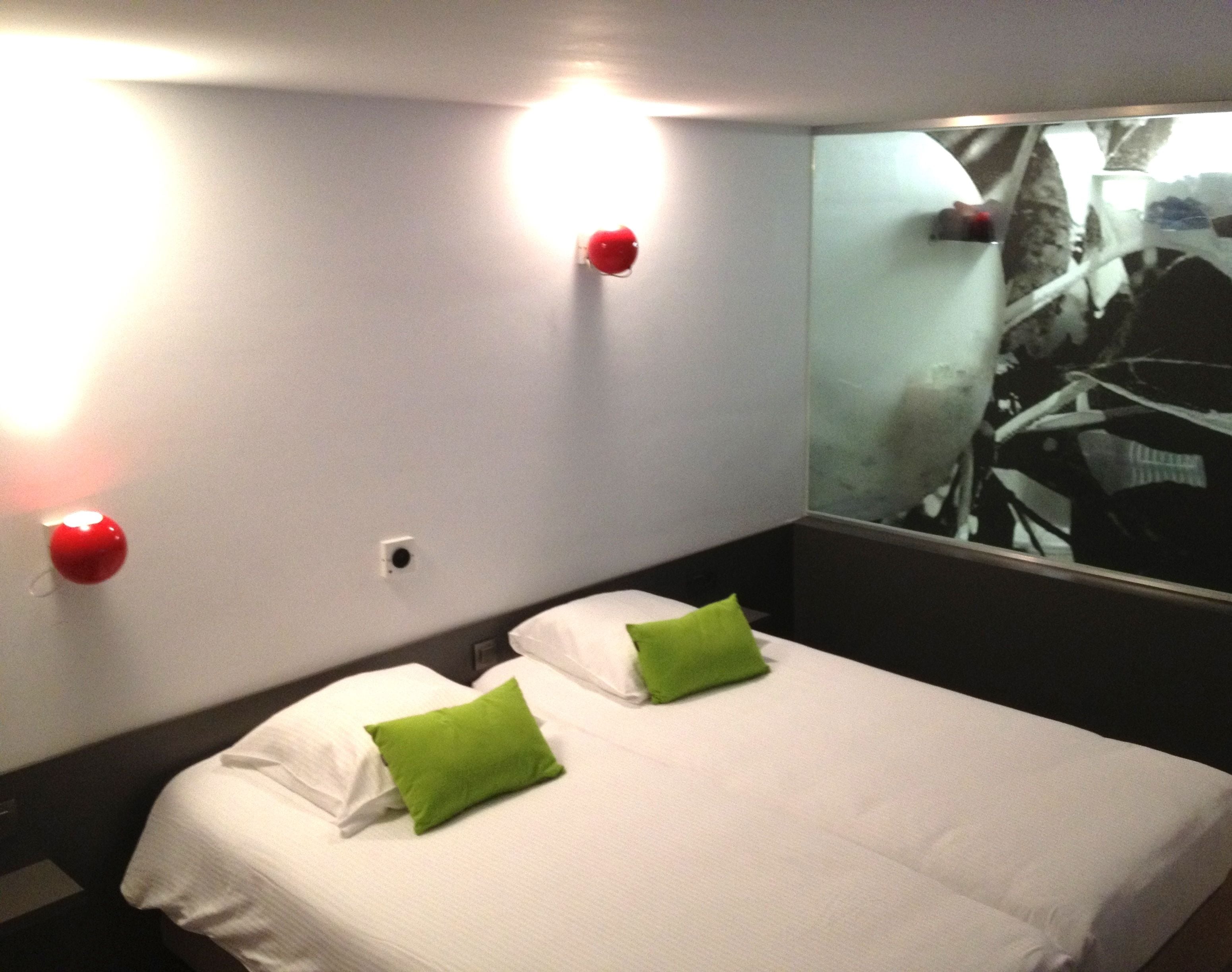 Quarto de hotel com cama, roupa de cama branca, almofadas verdes, um espelho e decorações vermelhas nas paredes.
