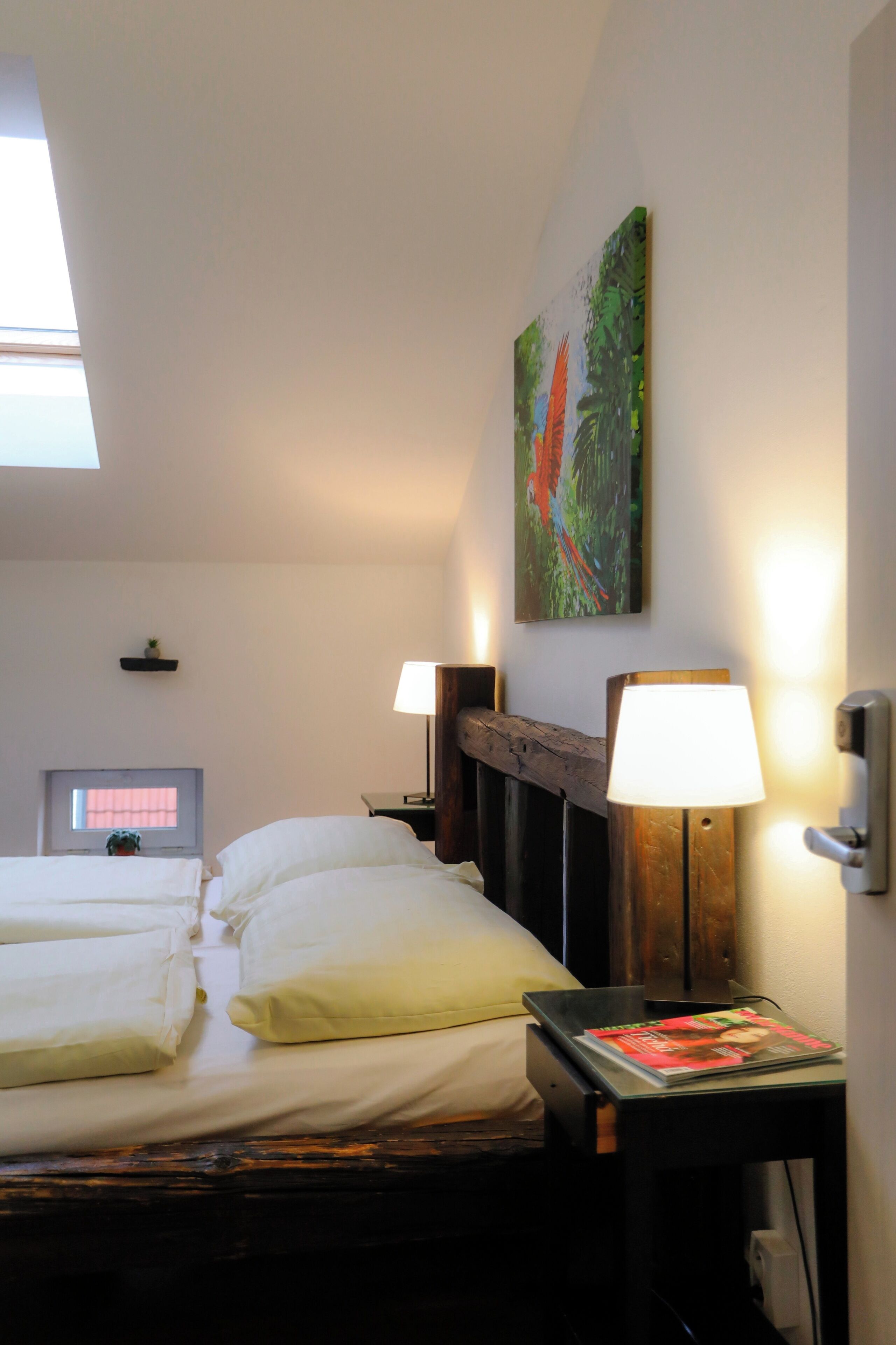 Quarto de hotel com duas camas, uma janela, um quadro, um abajur e uma revista na mesa de cabeceira.