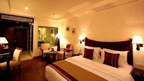 Quarto de hotel com uma cama grande, duas poltronas, uma mesinha, uma escrivaninha com abajur e um espelho.
