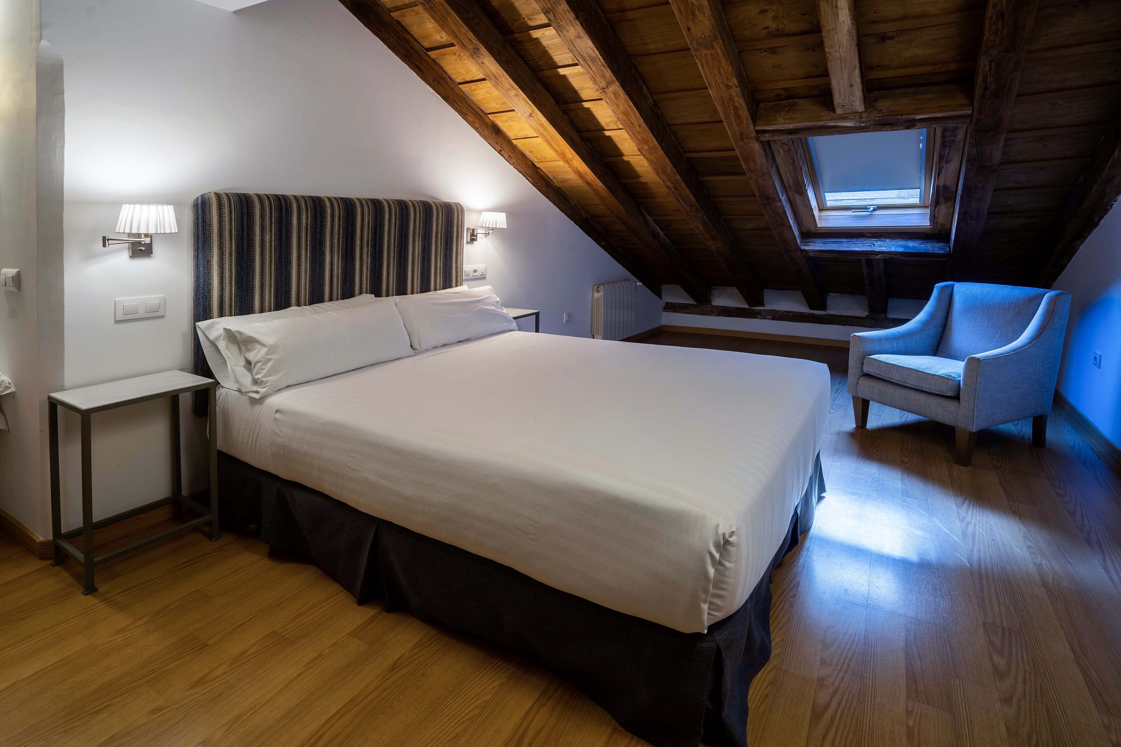 Quarto com cama, mesas de cabeceira, uma cadeira, claraboia e teto de madeira.