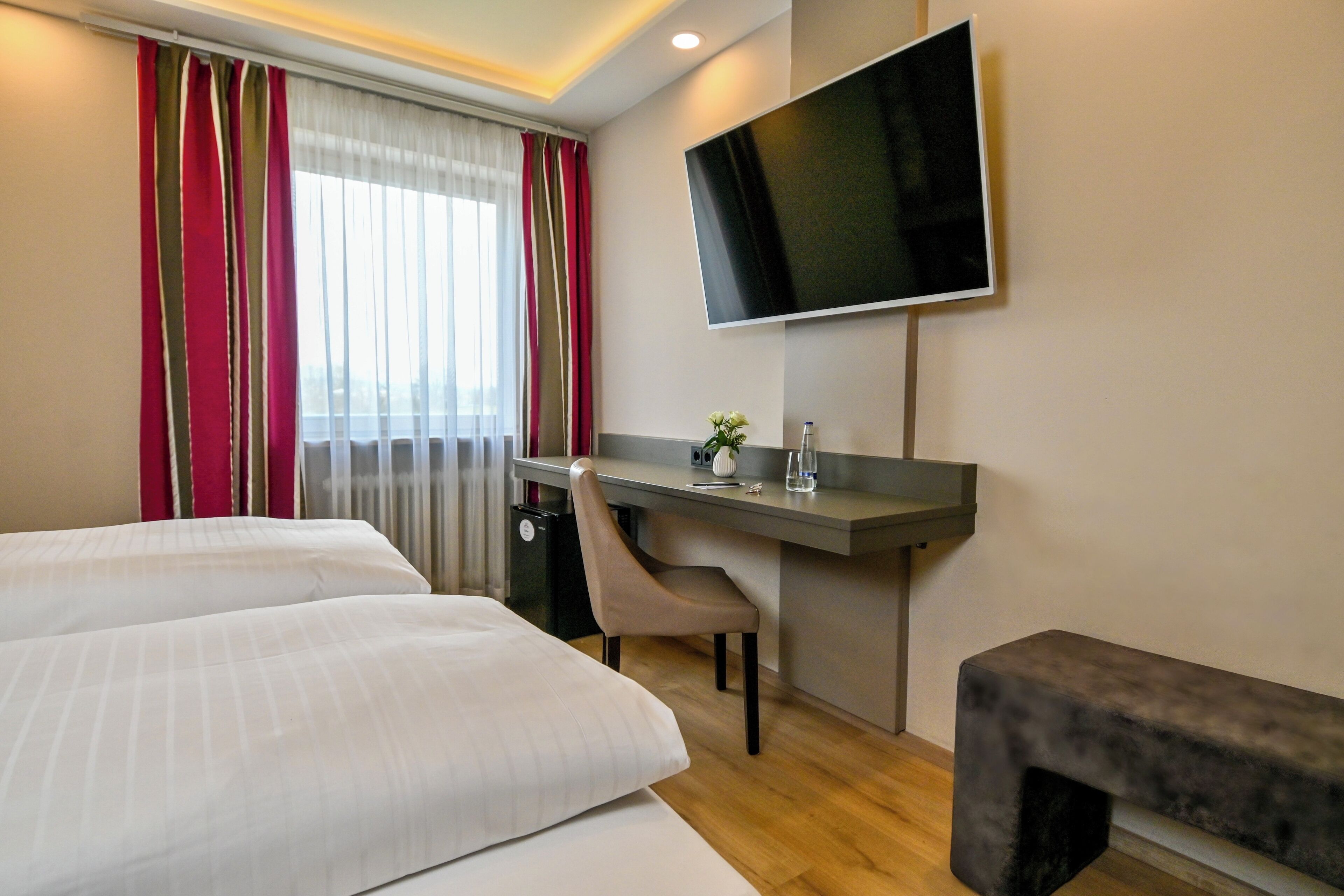 Quarto de hotel com duas camas, uma escrivaninha com cadeira, televisão na parede e um banquinho.