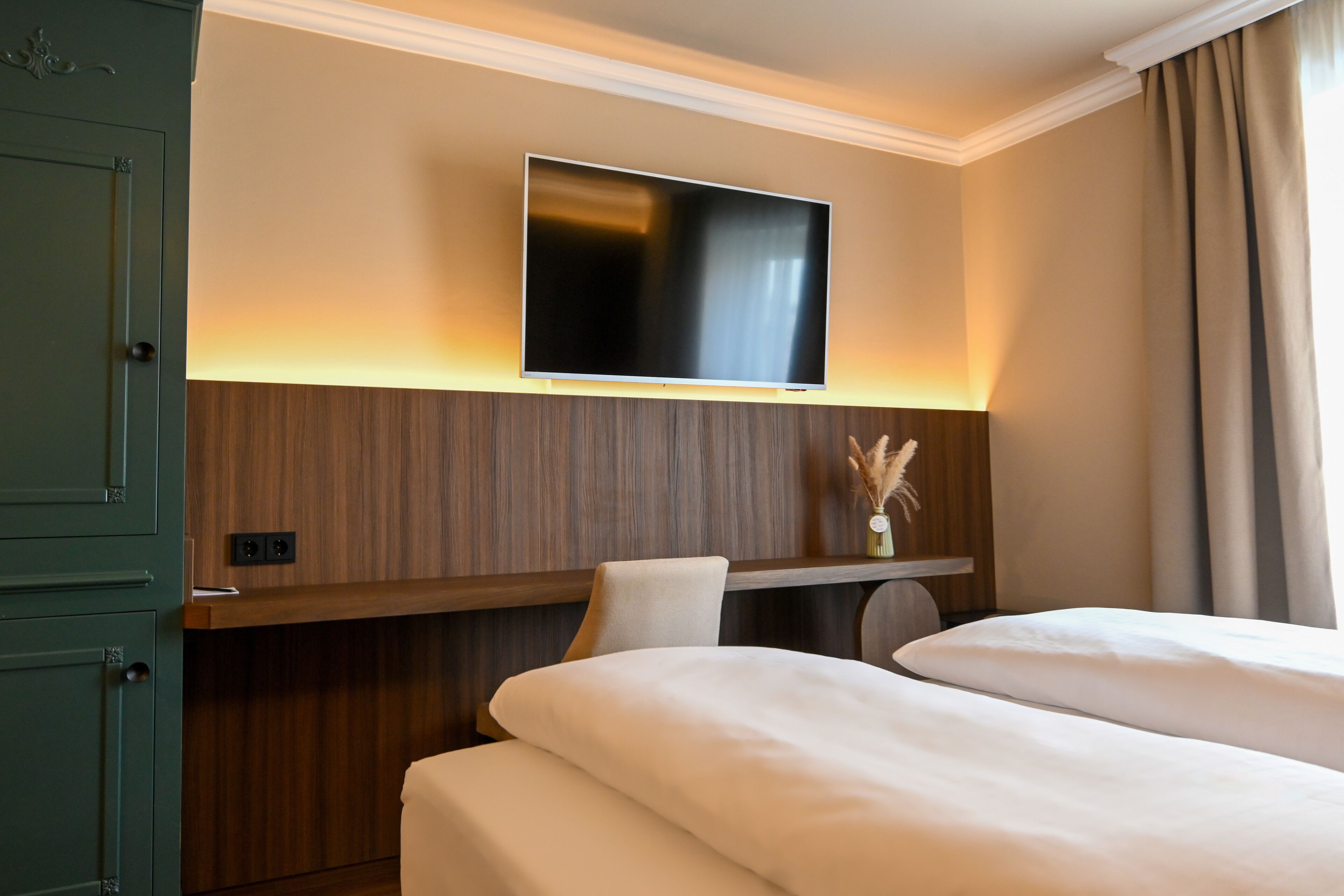 Quarto de hotel com televisão de tela plana na parede, painel de madeira, escrivaninha com cadeira e duas camas.