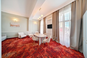 Quarto de hotel com carpete vermelho, mesa redonda com cadeiras, sofá, televisão e uma grande janela com cortinas.