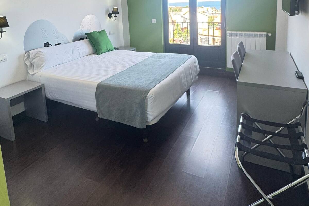 Quarto de hotel moderno com cama, mesas de cabeceira, escrivaninha e uma cadeira. Há uma janela com vista para prédios e uma varanda.