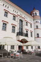 Um edifício com fachada branca, detalhes vermelhos e uma torre com telhado vermelho. Há mesas e cadeiras ao ar livre sob guarda-sóis.