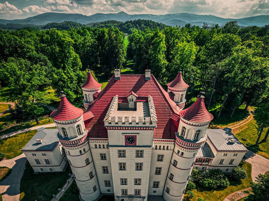 Um castelo com várias torres e uma entrada central, rodeado por vegetação exuberante e um céu limpo.