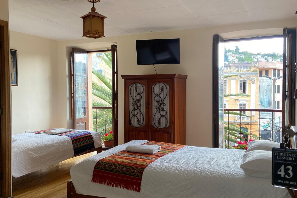 Quarto de hotel com duas camas, um armário de madeira, uma televisão e vista para prédios por uma janela grande.