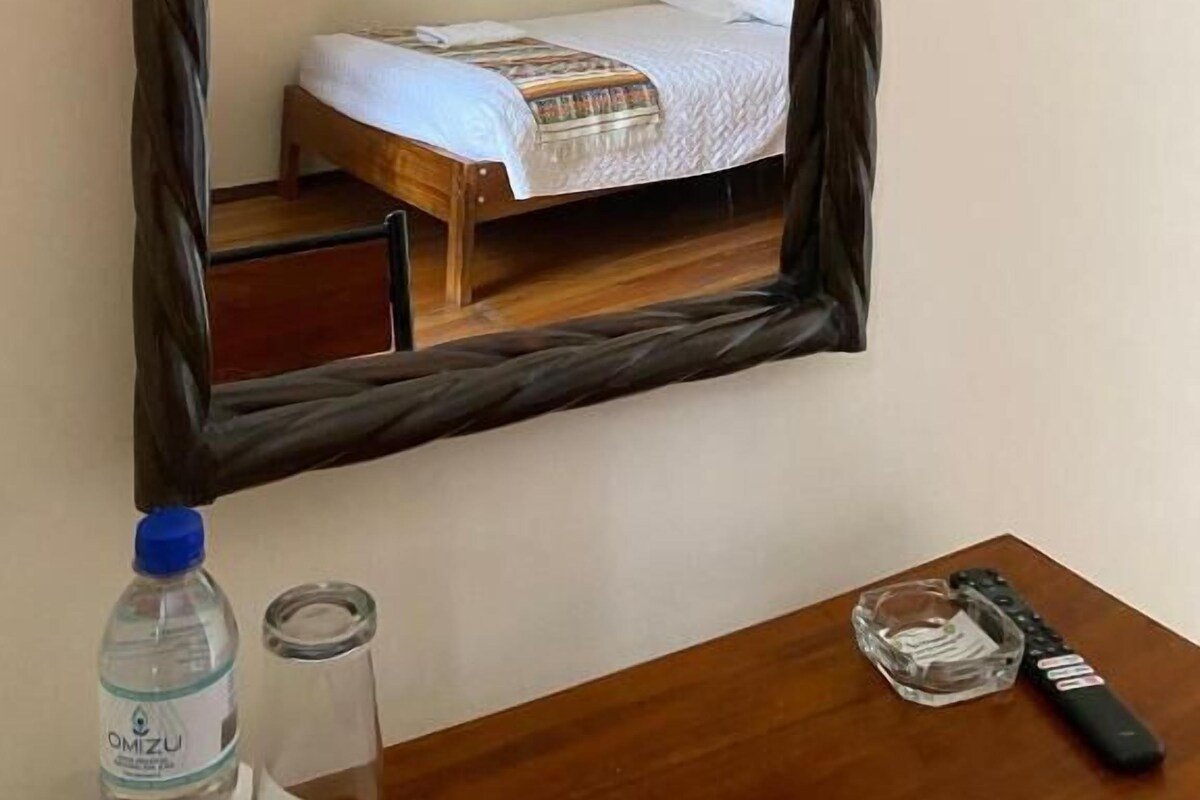 Quarto com uma cama, um espelho, uma mesa de madeira com um controle remoto, uma garrafa de água e um copo.