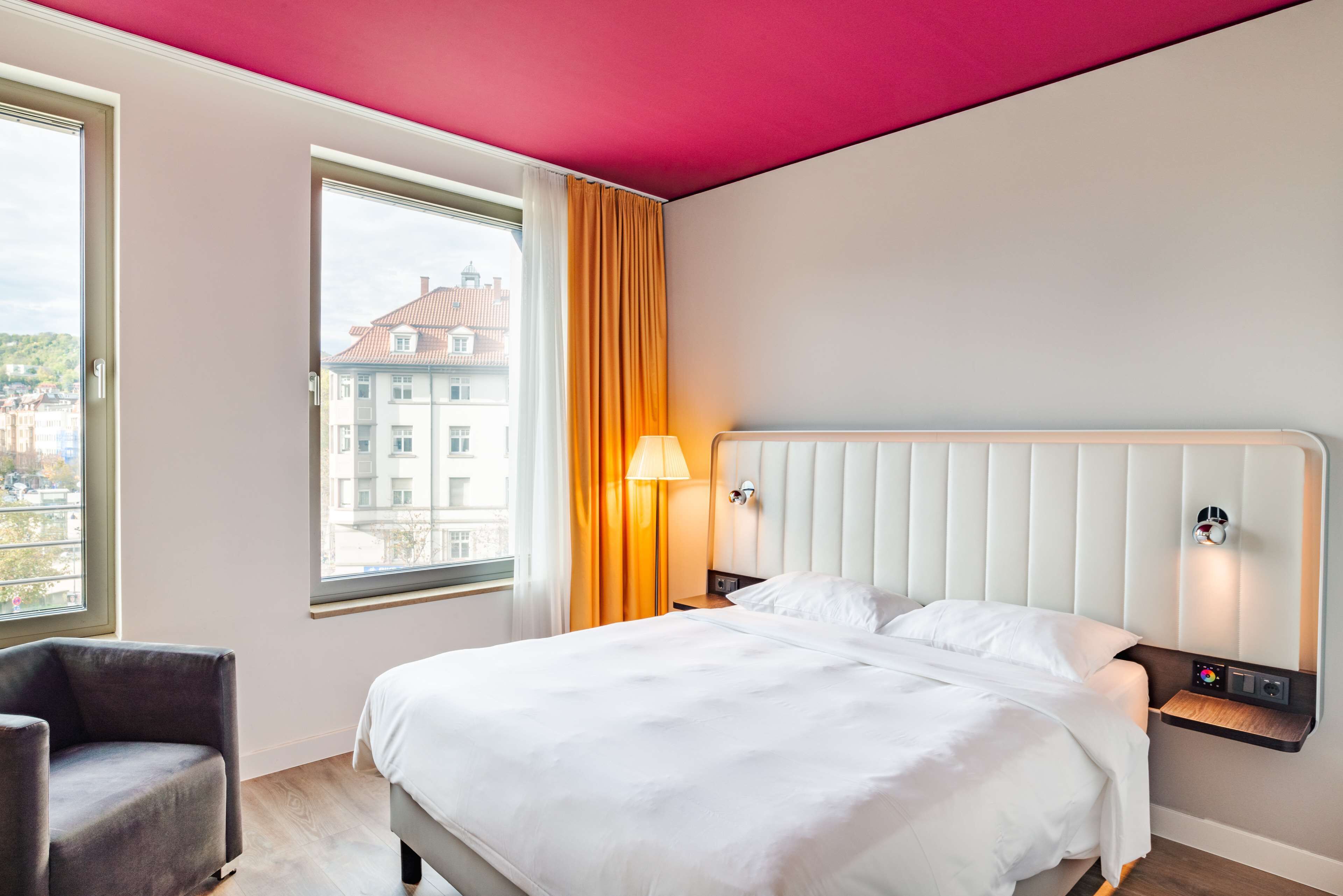 Quarto de hotel com uma cama grande, mesa de cabeceira, uma cadeira, janela com cortinas e vista para a cidade.