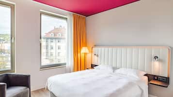 Quarto de hotel com uma cama grande, mesa de cabeceira, uma cadeira, janela com cortinas e vista para a cidade.