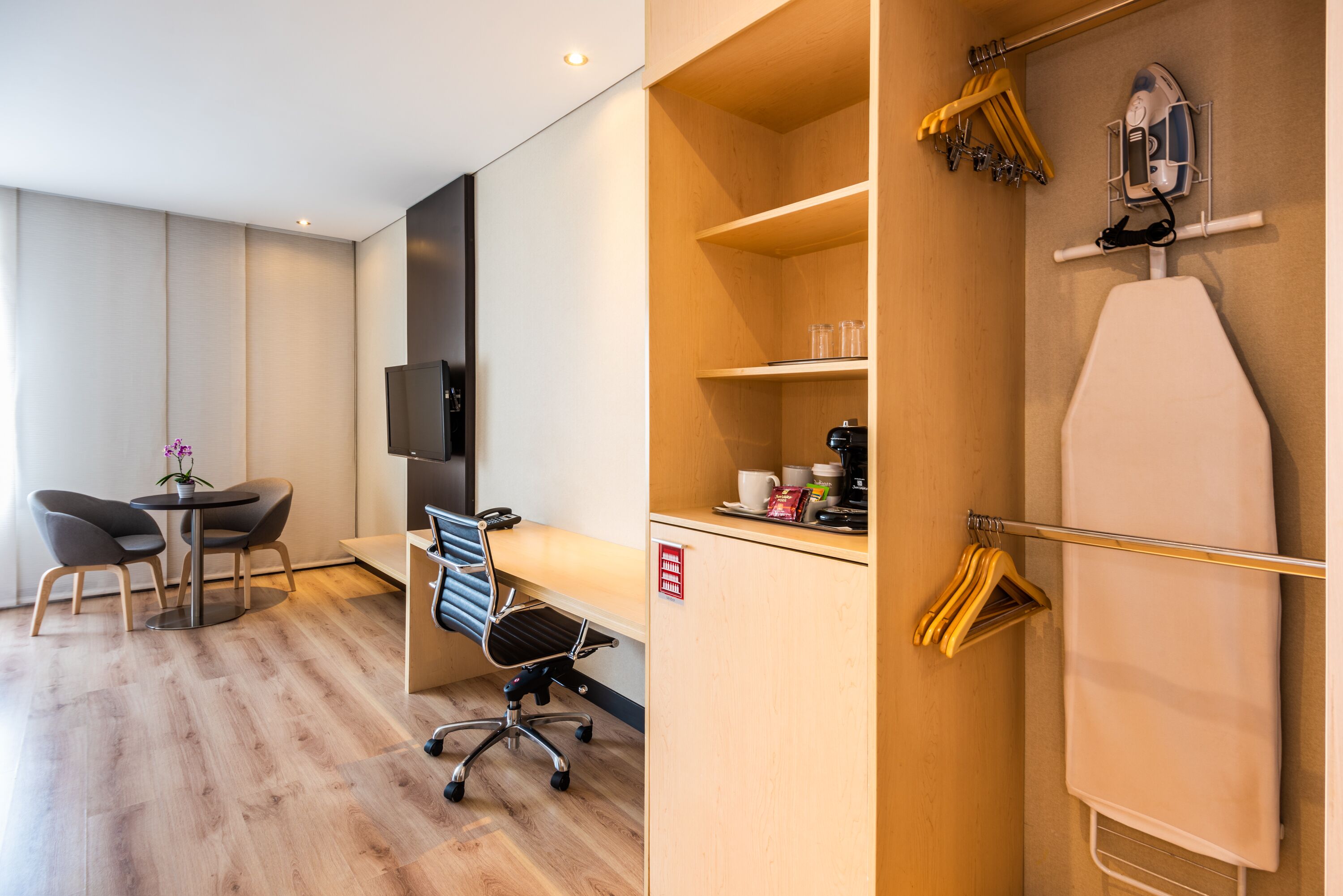 Quarto de hotel com piso de madeira, uma escrivaninha com cadeira, televisão, uma mesinha com planta em vaso e área de chuveiro com porta-toalhas.