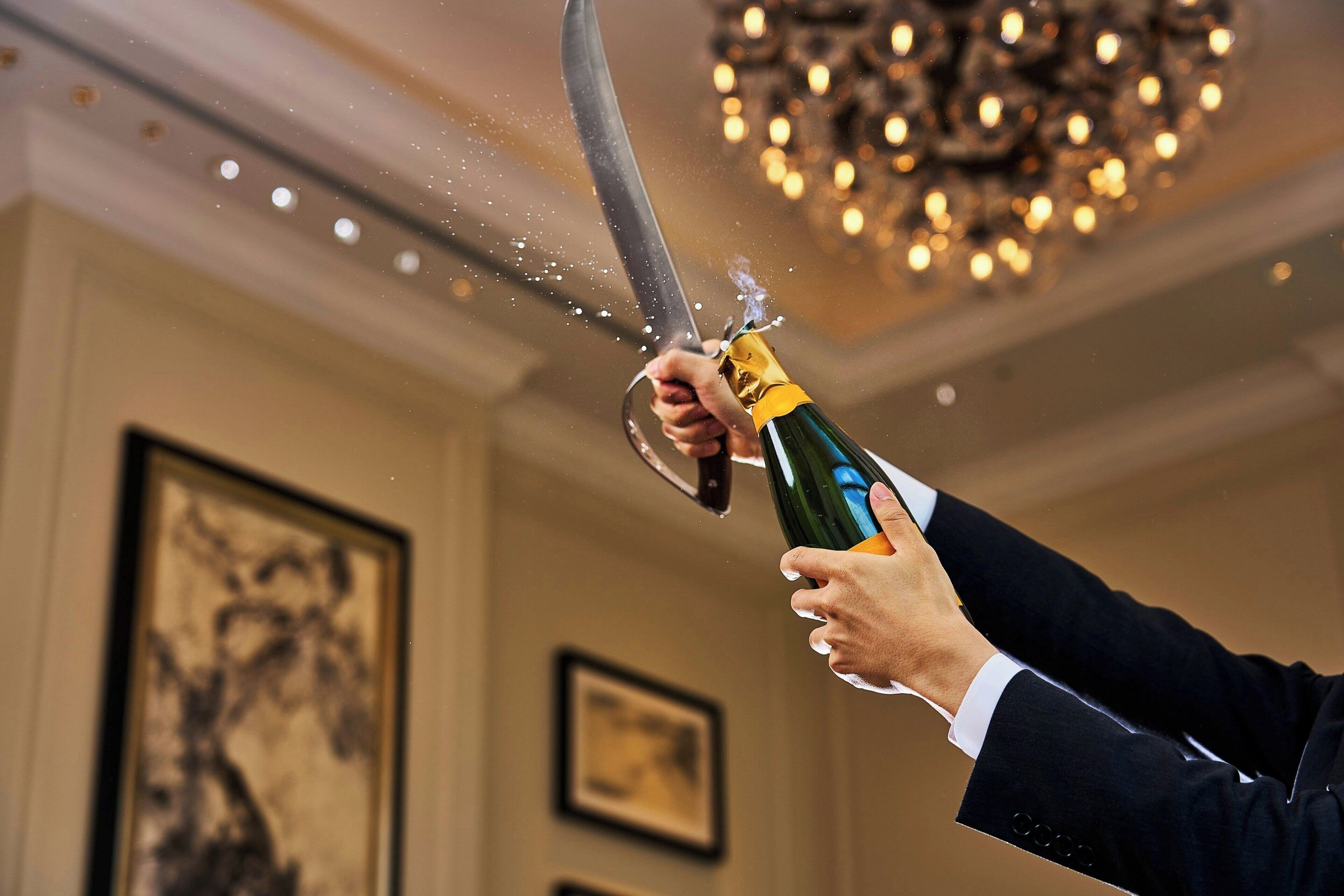 Uma pessoa segurando uma garrafa de champanhe com uma espada, com um candelabro ao fundo.