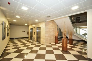 Um saguão de hotel com piso xadrez, elevadores, escadas e plantas decorativas.
