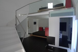Quarto de hotel moderno com escada, uma cama, uma pequena escrivaninha e um banheiro visível por uma porta aberta.