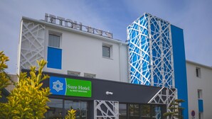 Um hotel moderno com fachada azul e branca marcante, uma placa com a inscrição 'Sure Hotel by Best Western' e uma câmera de segurança.