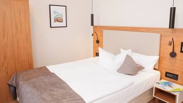 Cama bem arrumada com lençóis brancos e manta marrom decorativa, cabeceira de madeira, mesinha de cabeceira com uma revista e um quadro na parede.