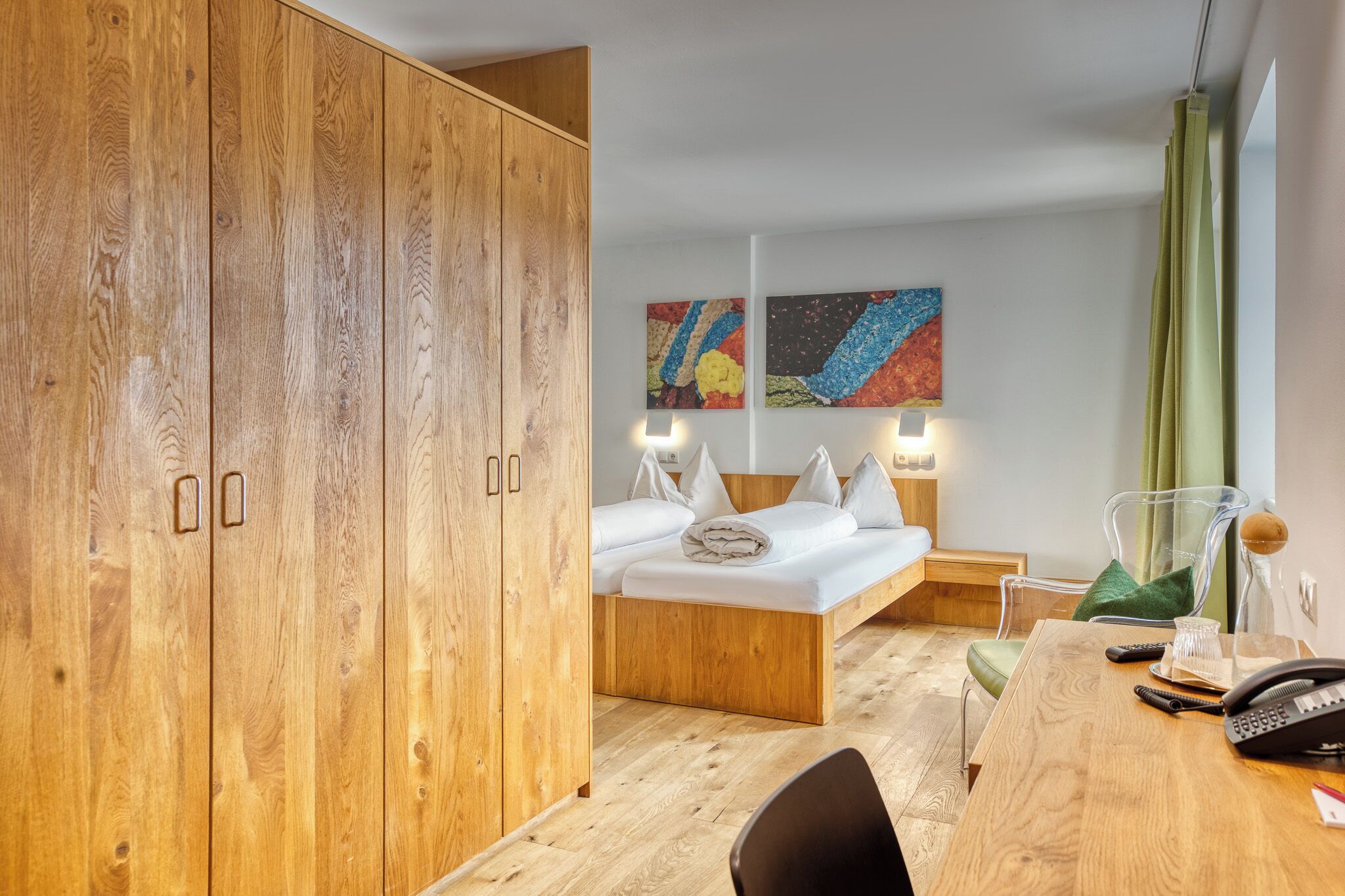 Quarto moderno com guarda-roupa de madeira, cama com roupa de cama branca, escrivaninha com cadeira e duas luminárias de parede.