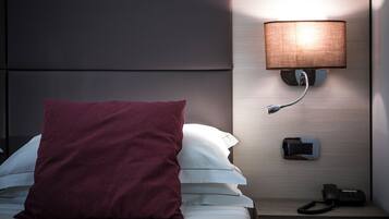 Cama bem arrumada com lençóis brancos e uma única almofada vermelha, mesa de cabeceira com abajur, telefone e tomada.