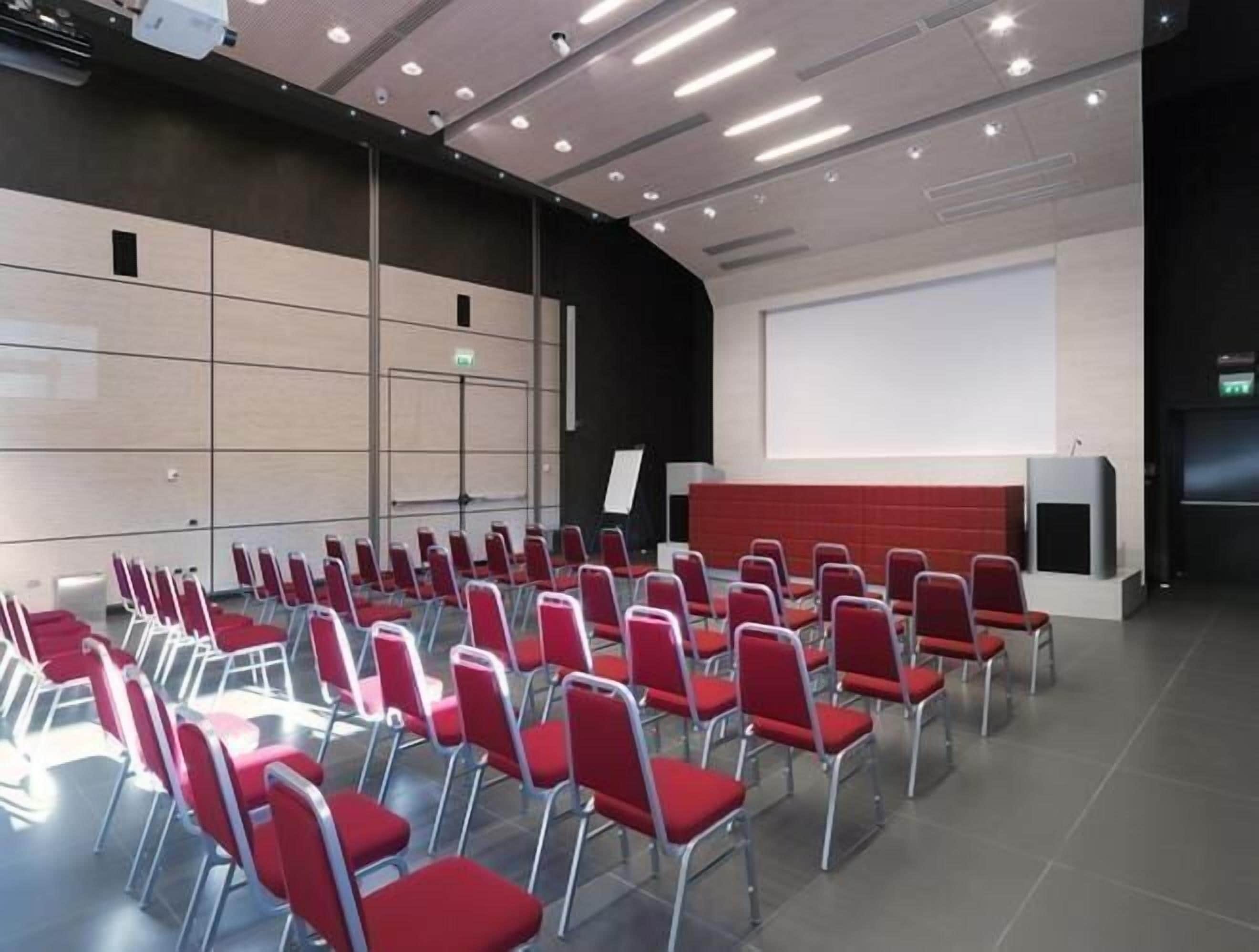 Uma sala de conferências com fileiras de cadeiras vermelhas, uma tela grande e um pódio.