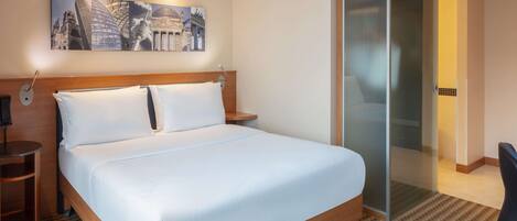 Quarto de hotel com uma cama grande, uma mesinha de cabeceira, uma escrivaninha, uma cadeira, uma porta de vidro e uma pintura com vista para a cidade na parede.