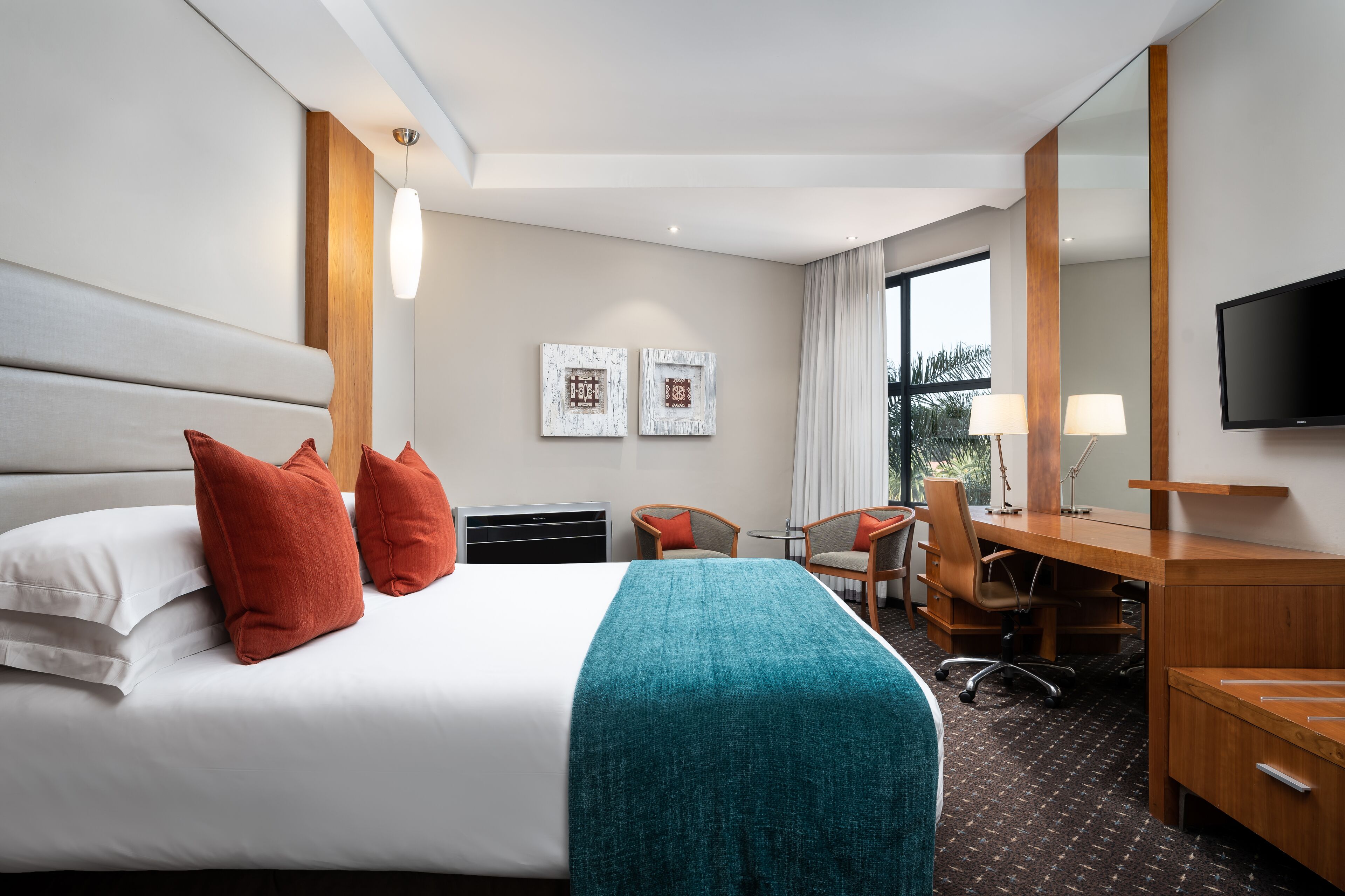 Quarto de hotel com uma cama grande, uma escrivaninha, uma cadeira, uma televisão e duas molduras na parede.