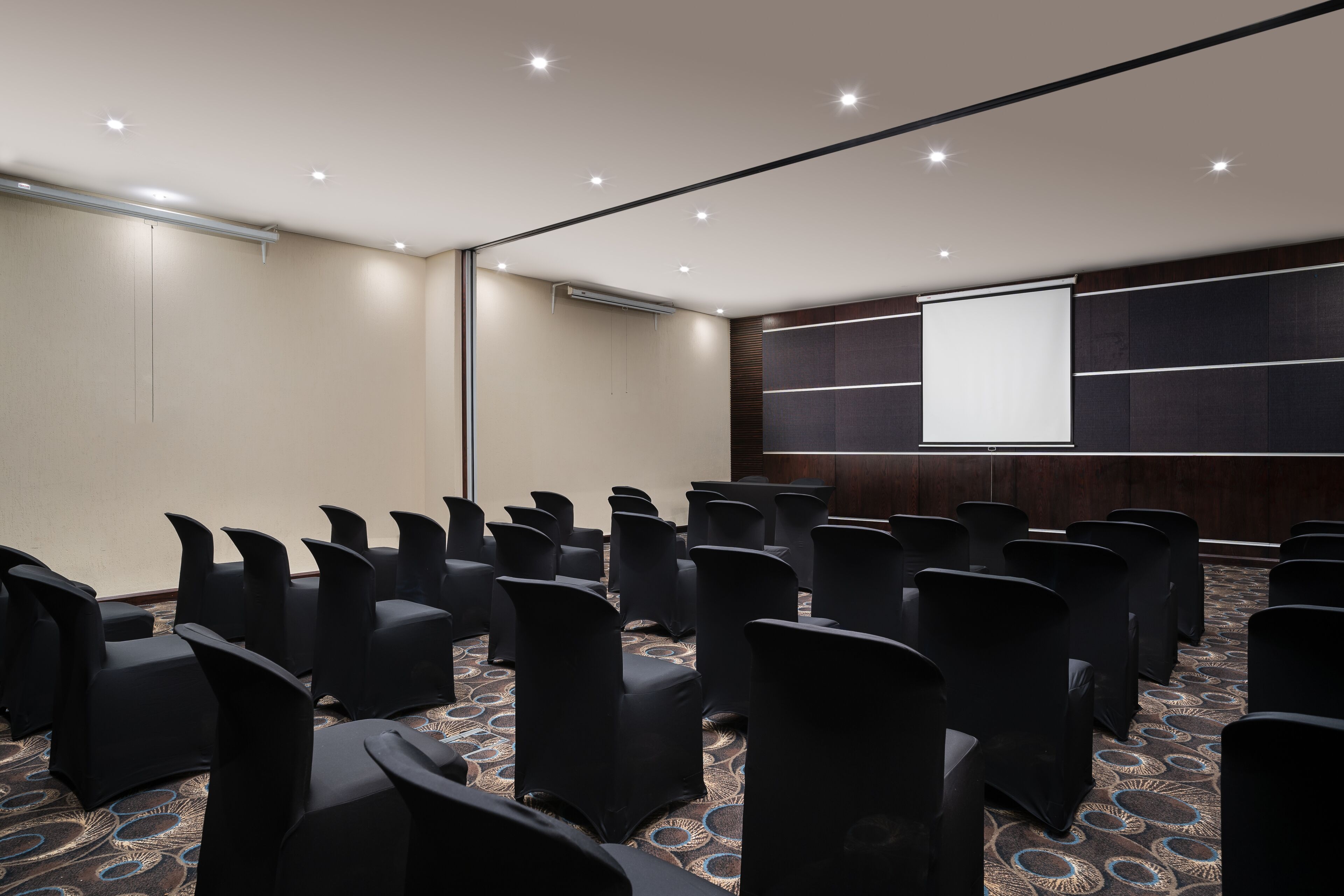 Uma sala de conferências com fileiras de cadeiras pretas, uma tela de projetor e piso coberto de carpete.