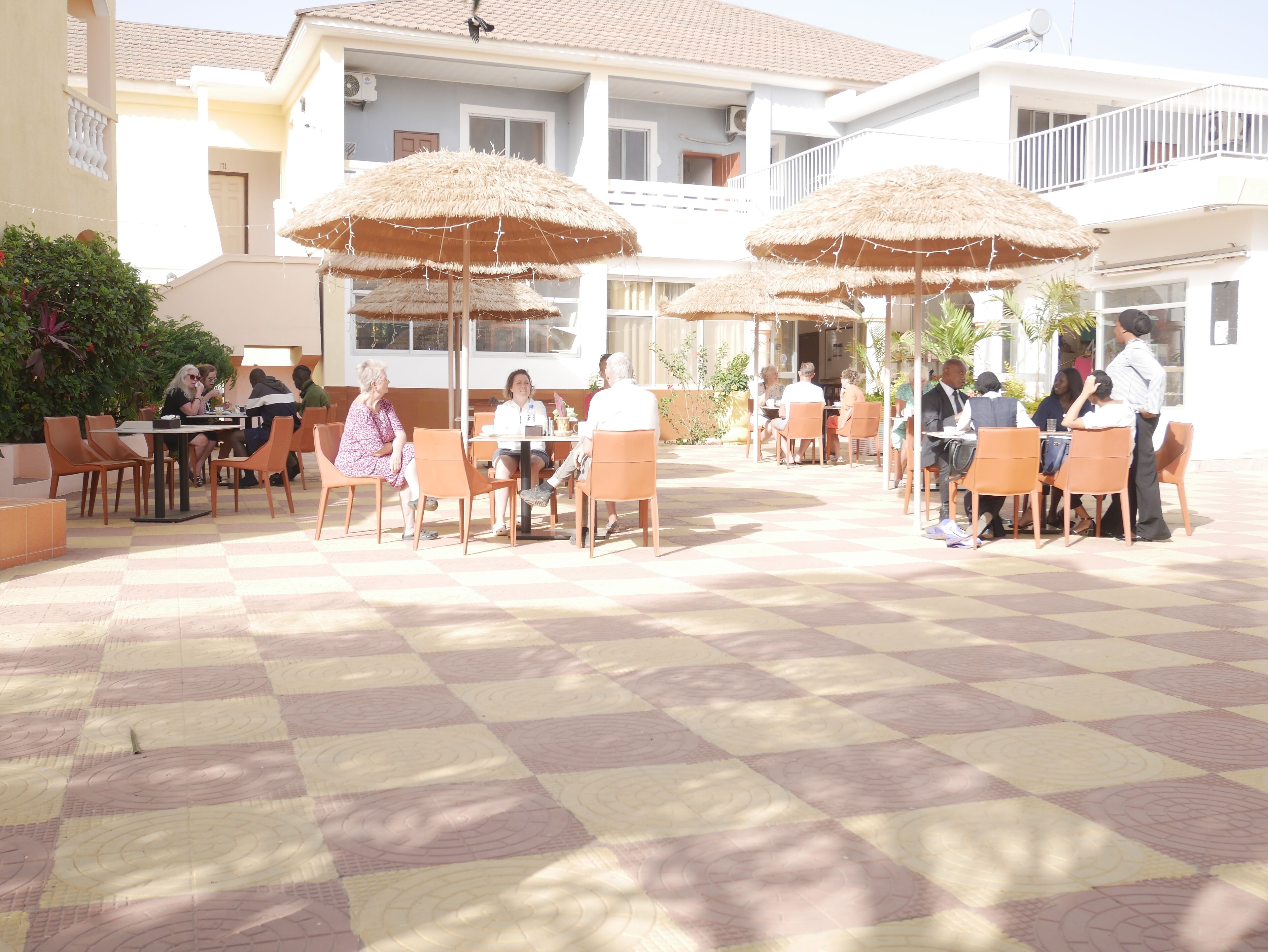 Uma área de pátio ao ar livre com guarda-sóis de palha, mesas e cadeiras externas, e um padrão de piso quadriculado.