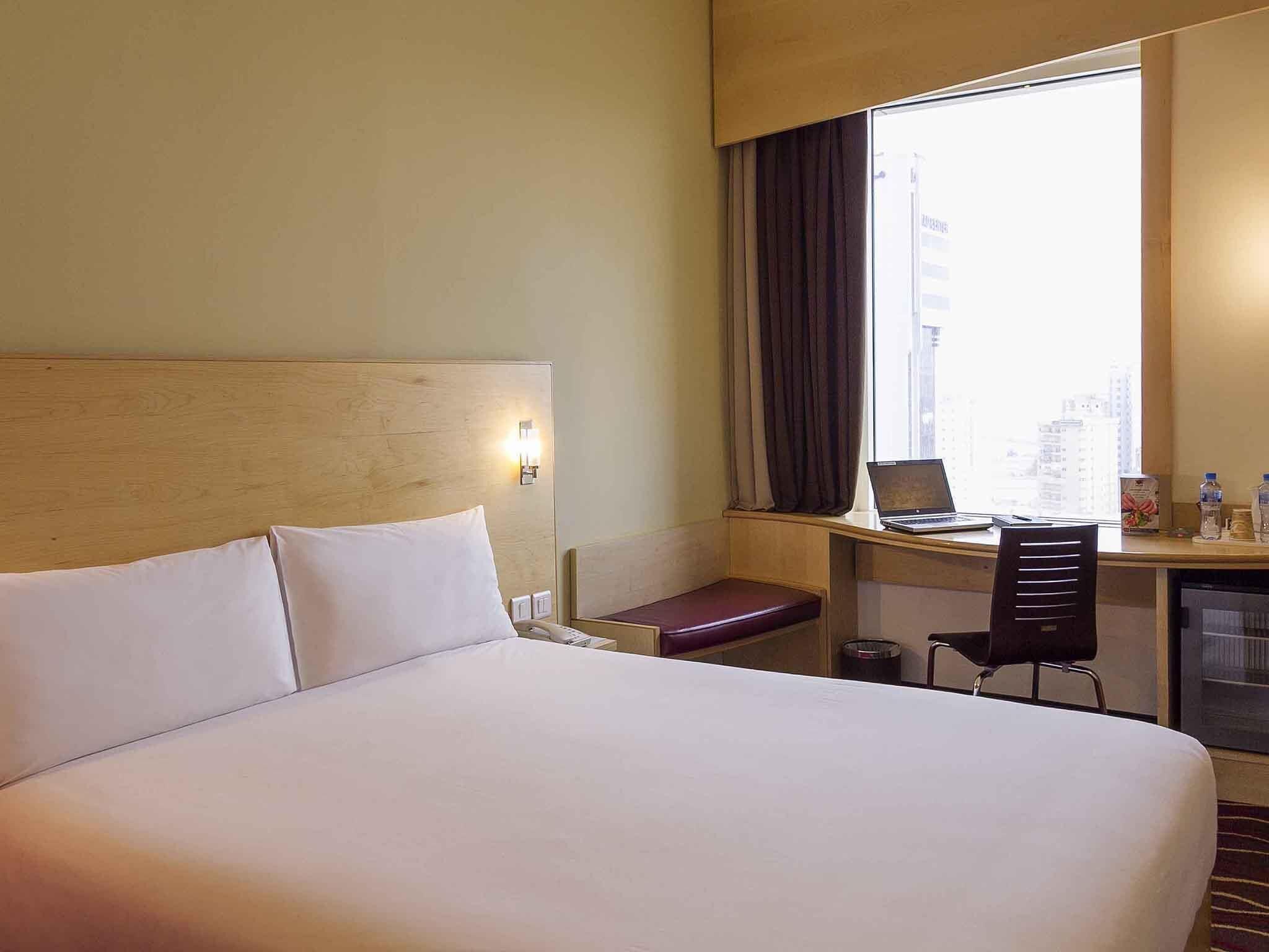 Quarto de hotel com uma cama grande, uma escrivaninha com um laptop, uma cadeira e uma janela com cortinas.