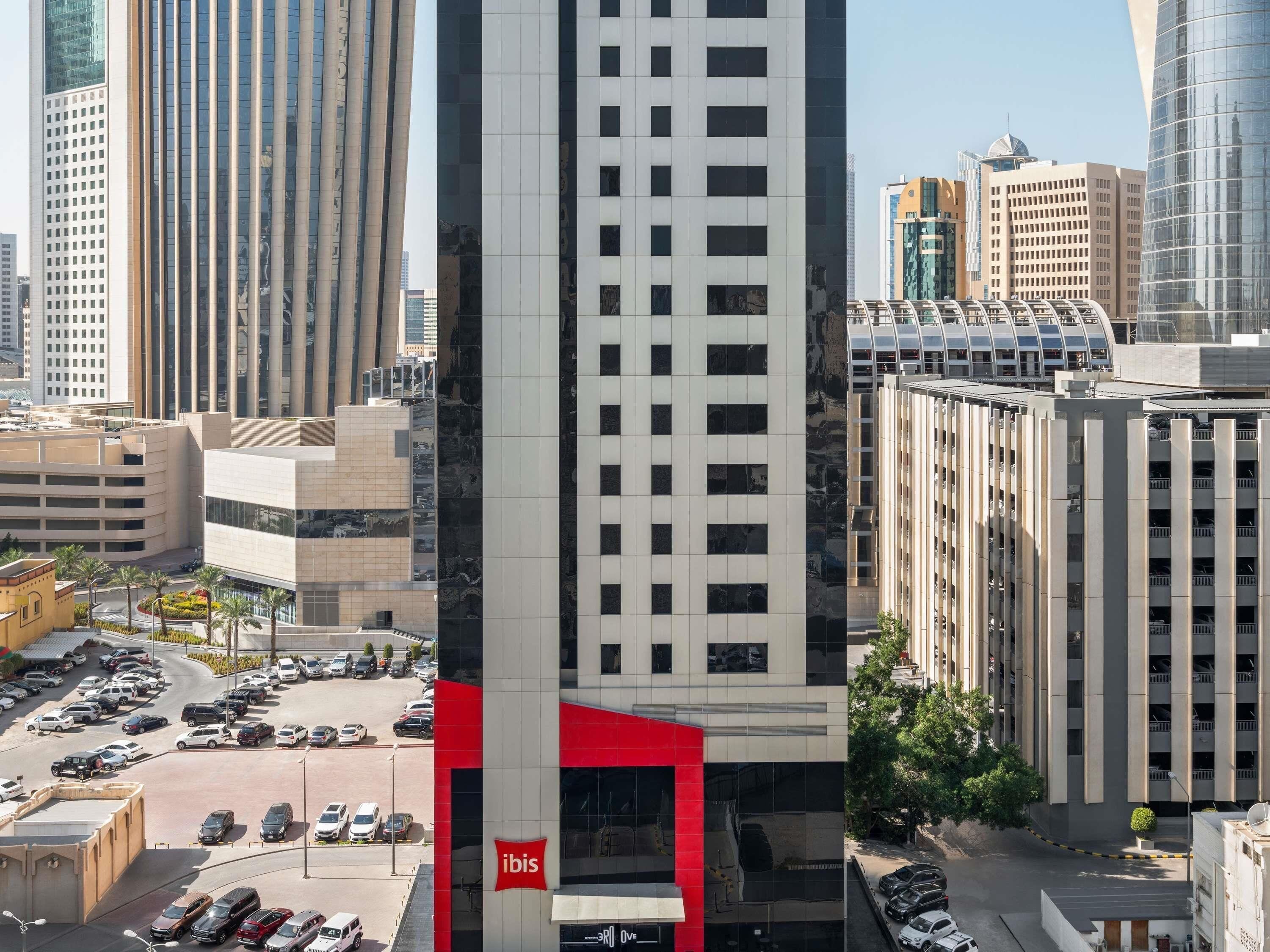 Um edifício alto com fachada marcante em vermelho e branco, cercado por outros edifícios modernos e um estacionamento com vários carros.