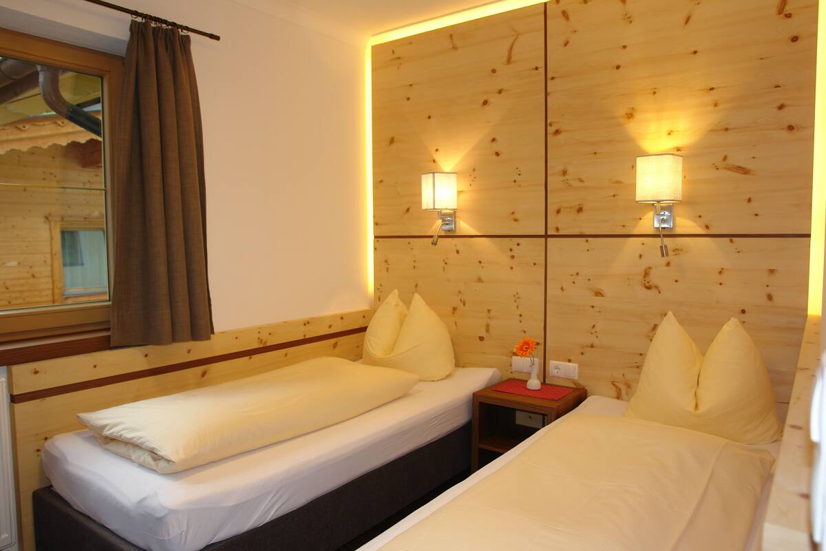 Quarto de hotel com duas camas, cabeceira de madeira, mesa de cabeceira com abajur e janela com cortinas.