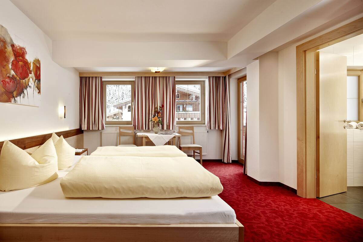 Quarto de hotel com uma cama grande, uma mesa de jantar com cadeiras e vista para um edifício pelas janelas.