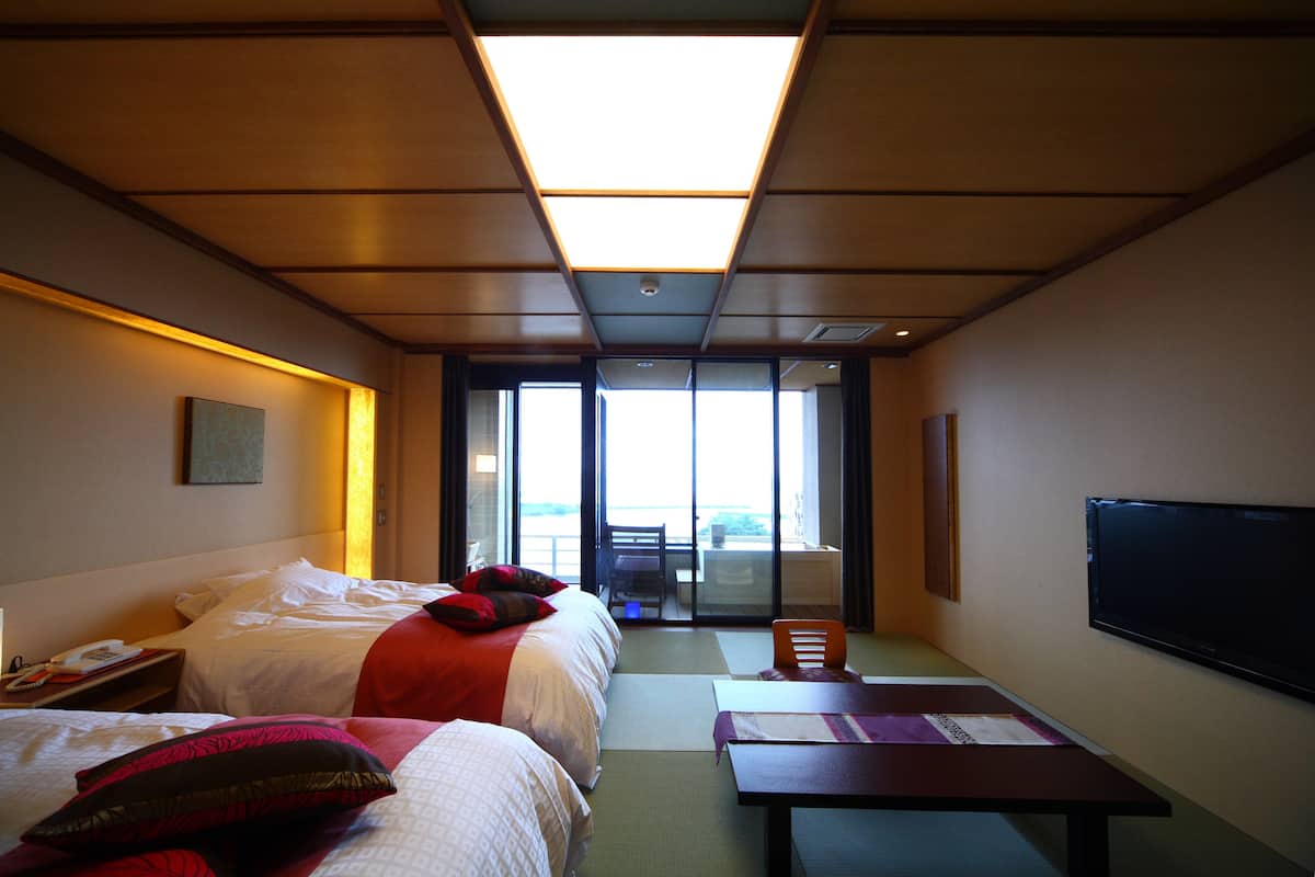 Quarto de hotel com duas camas, uma mesa, uma cadeira, televisão e vista para o exterior através de uma porta de correr.